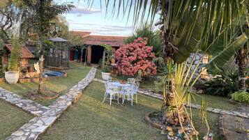 Um jardim com um caminho de pedra, uma mesa e cadeiras brancas, e diversas plantas e árvores.