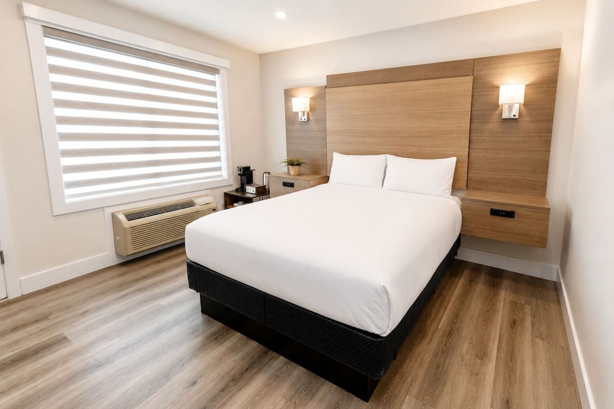 Quarto de hotel moderno com uma cama grande, cabeceira embutida, abajures e uma janela com persianas.