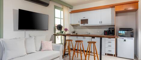 Cozinha moderna com armários brancos, micro-ondas embutido e cafeteira. Há uma área de jantar com mesa de madeira e quatro banquetas. Uma TV de tela plana está fixada na parede acima do sofá.