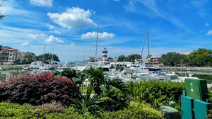 Uma marina com vários barcos atracados, um farol ao fundo e vegetação exuberante em primeiro plano.