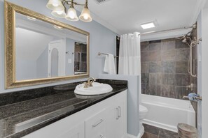 Casa de banho com bancada de mármore, espelho com moldura dourada, lavatório branco, banheira e chuveiro com cortina.