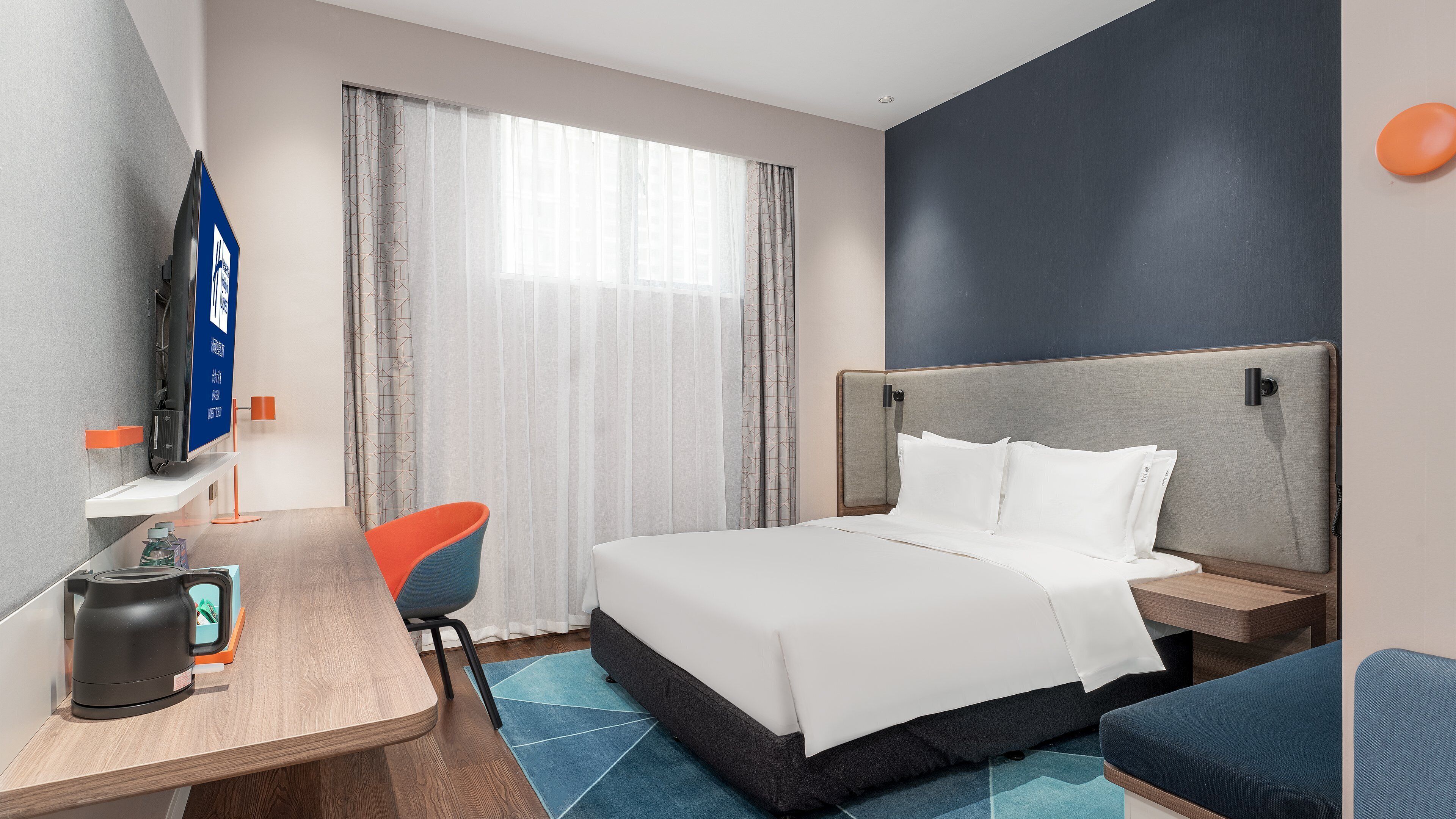 Quarto de hotel com uma cama, uma escrivaninha com cadeira, televisão e janela com cortinas.