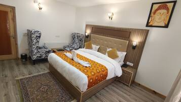 Quarto de hotel com uma cama grande, duas poltronas, uma mesa pequena, uma mesinha de cabeceira, um ventilador de teto e um quadro de Buda na parede.