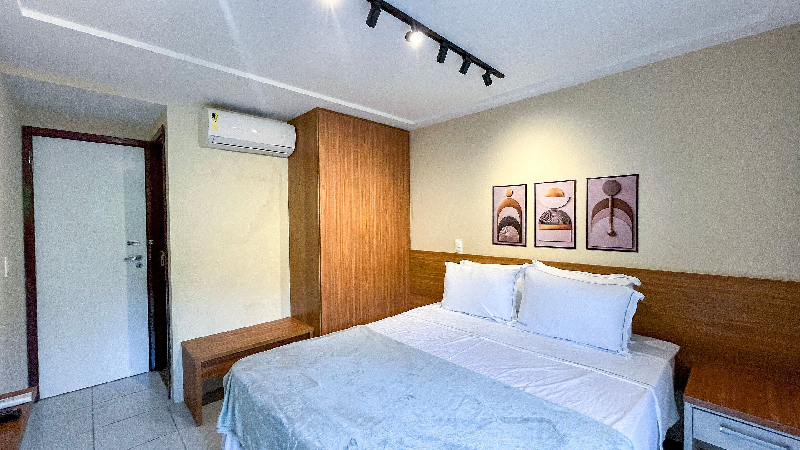 Quarto de hotel com cama, cabeceira de madeira, um banco, guarda-roupa e três obras de arte penduradas na parede.