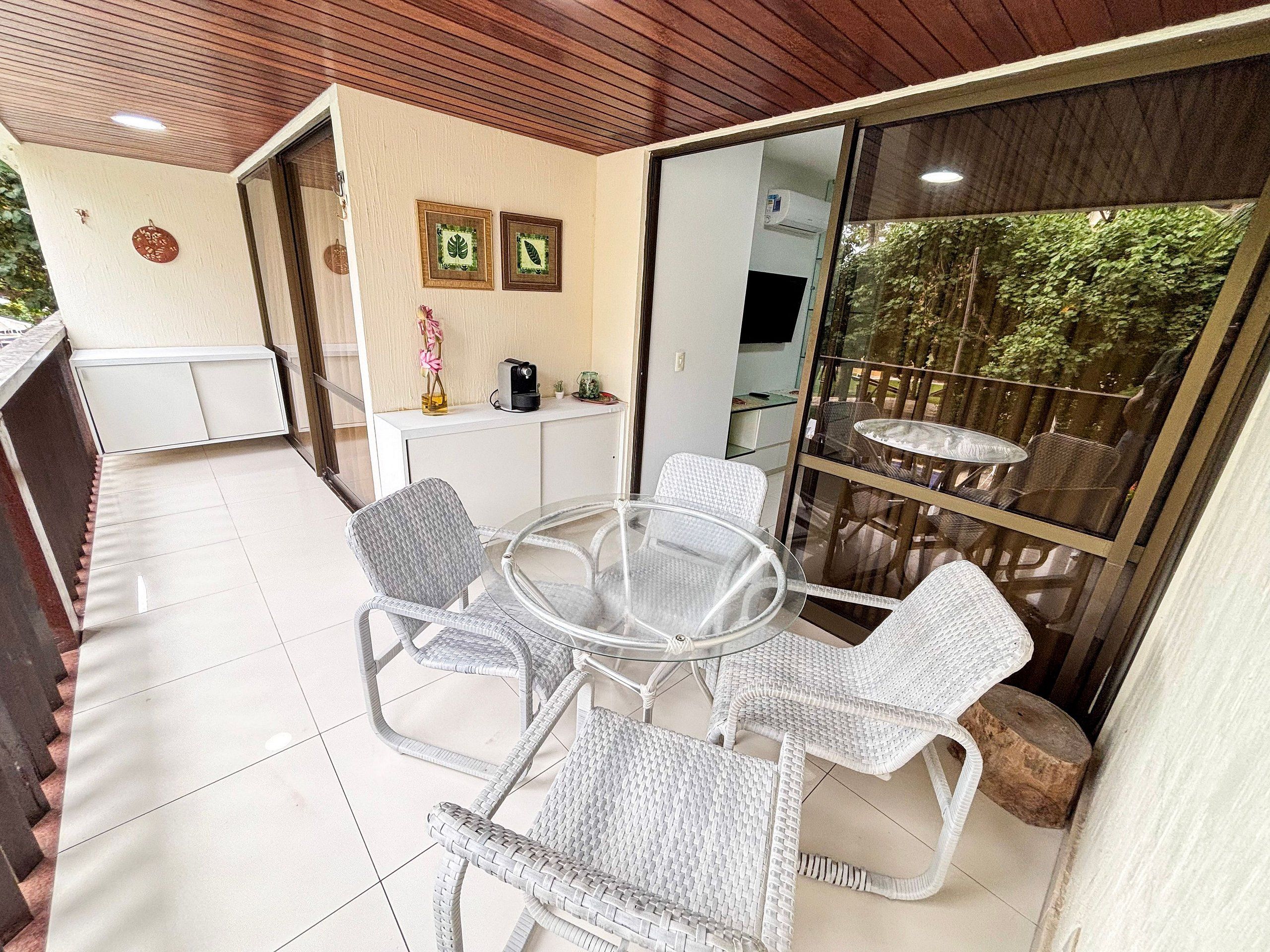 Uma área externa coberta com mesa de vidro e cadeiras de vime, um pequeno espaço de cozinha com geladeira e cafeteira, e uma porta de vidro deslizante que dá para um jardim.