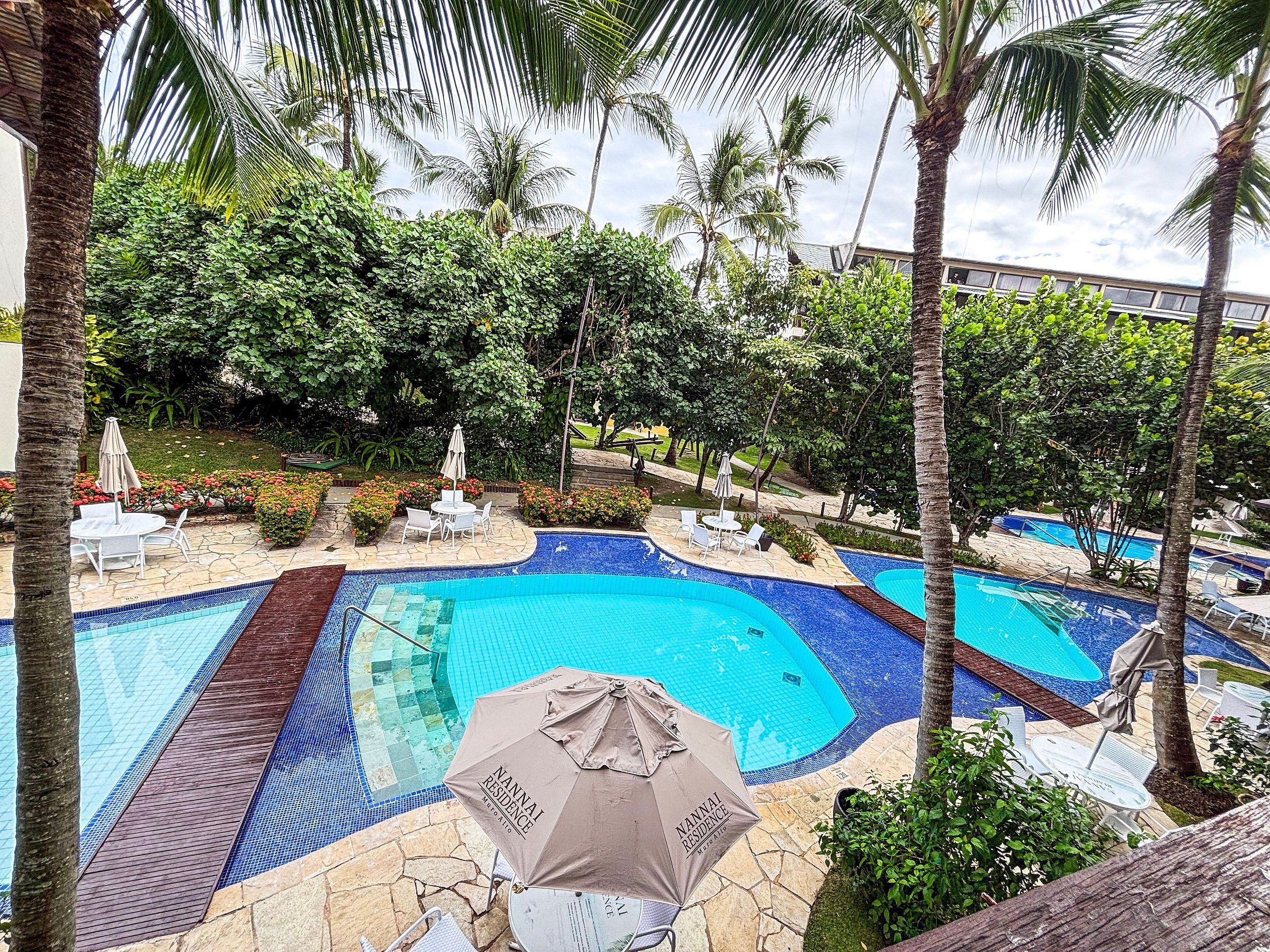 Área de piscina com espreguiçadeiras e guarda-sóis, cercada por palmeiras e vegetação.