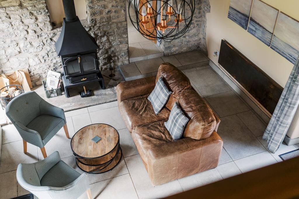 Uma sala de estar acolhedora com parede de pedra, uma lareira a lenha preta, um sofá de couro com almofadas e uma mesa de centro redonda de madeira.