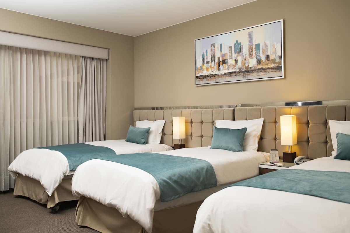 Quarto de hotel com três camas, uma pintura de paisagem urbana e uma janela grande com cortinas.