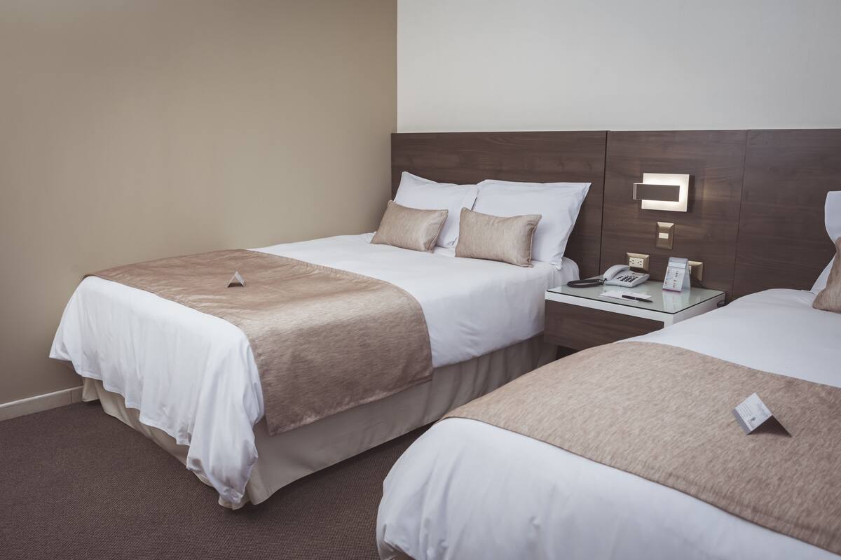 Quarto de hotel com duas camas, uma mesinha de cabeceira com telefone e abajur, e uma luminária na parede.