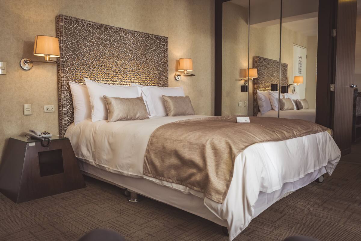 Quarto de hotel com uma cama grande, cabeceira com design estampado, duas luminárias fixadas na parede, mesa de cabeceira com telefone e um espelho na parede.
