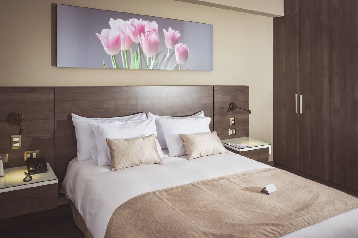 Quarto de hotel com uma cama grande, duas mesas de cabeceira, um guarda-roupa e um quadro na parede com tulipas cor-de-rosa.