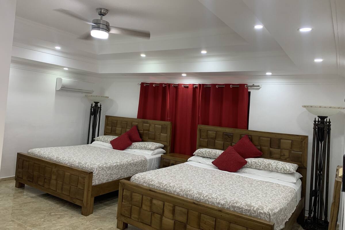 Quarto de hotel com duas camas de madeira, um ventilador de teto e uma cortina vermelha.