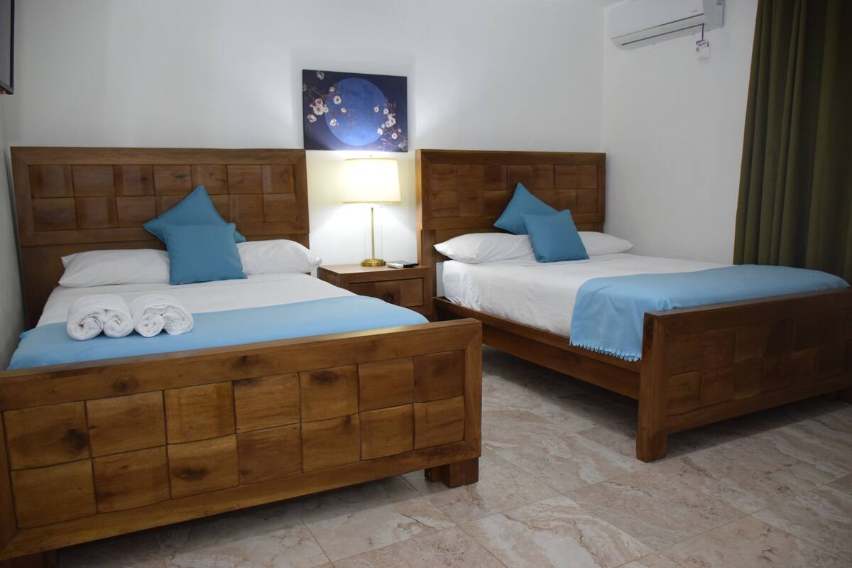 Duas camas de madeira com roupa de cama azul, uma mesa de cabeceira com abajur e um ar condicionado fixado na parede.