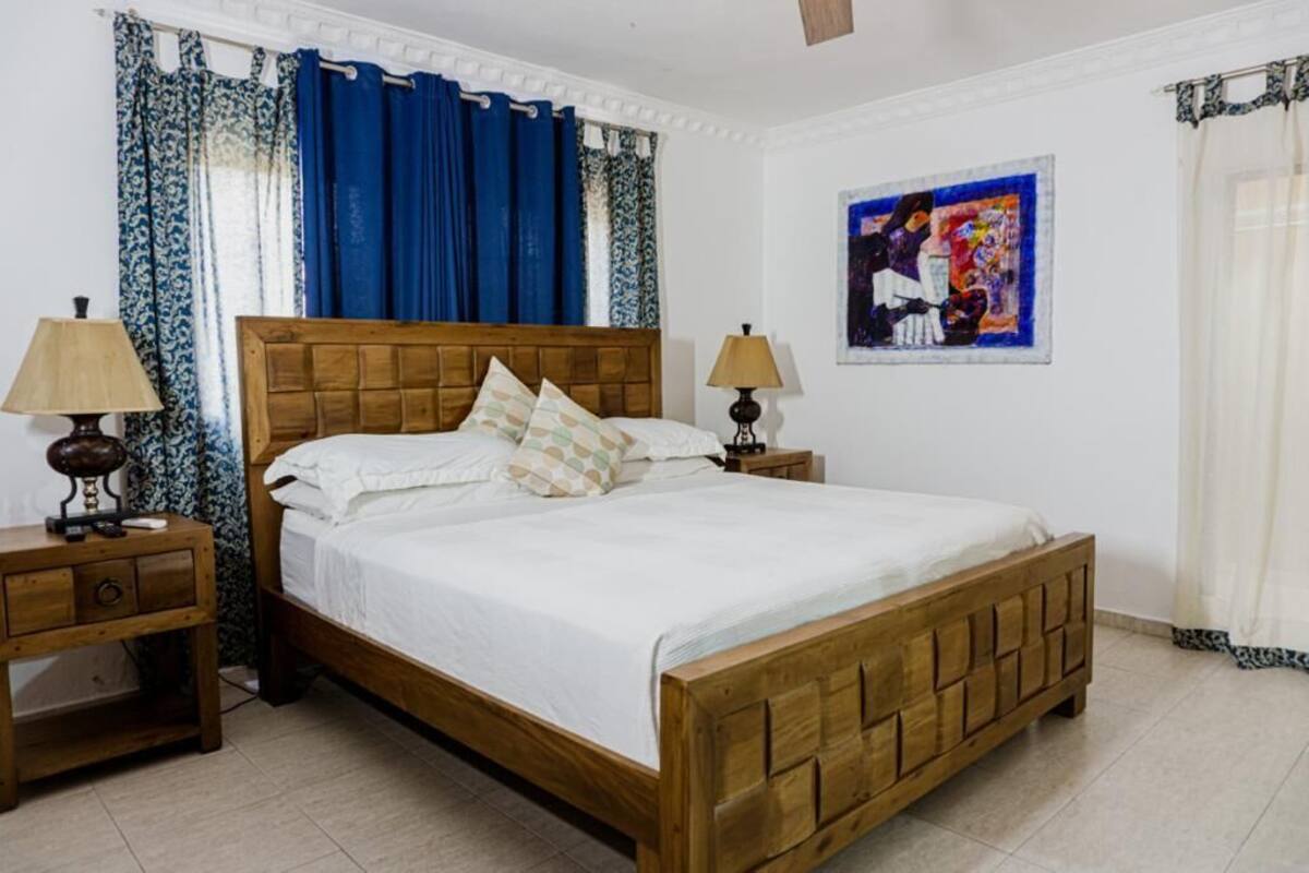 Quarto com cama de madeira, duas mesas de cabeceira com luminárias, cortinas estampadas e um quadro na parede.