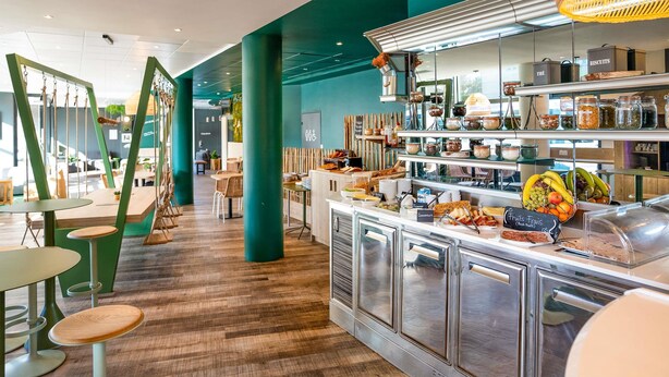 Um café moderno com piso de madeira, detalhes em verde e diversas opções de comida e bebida expostas em prateleiras.