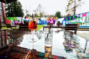 Uma mesa de vidro com uma bebida, bancos e um mural colorido ao fundo.
