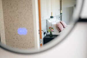 Casa de banho com espelho refletindo uma cama, mesa de cabeceira com um vaso de flores e um porta-toalhas.