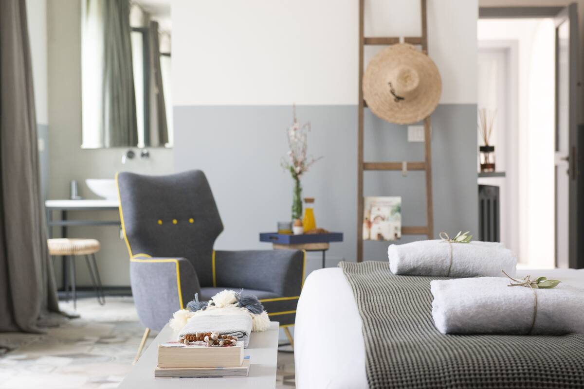 Quarto de hotel moderno com cama, uma cadeira, uma mesinha com um livro e objetos decorativos, um chapéu fixado na parede e vista para o corredor.