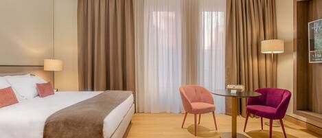 Quarto de hotel com uma cama, duas cadeiras, uma mesinha e um abajur.