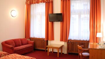 Quarto de hotel com sofá vermelho, uma pequena mesa de madeira, televisão fixada na parede e escrivaninha com abajur.