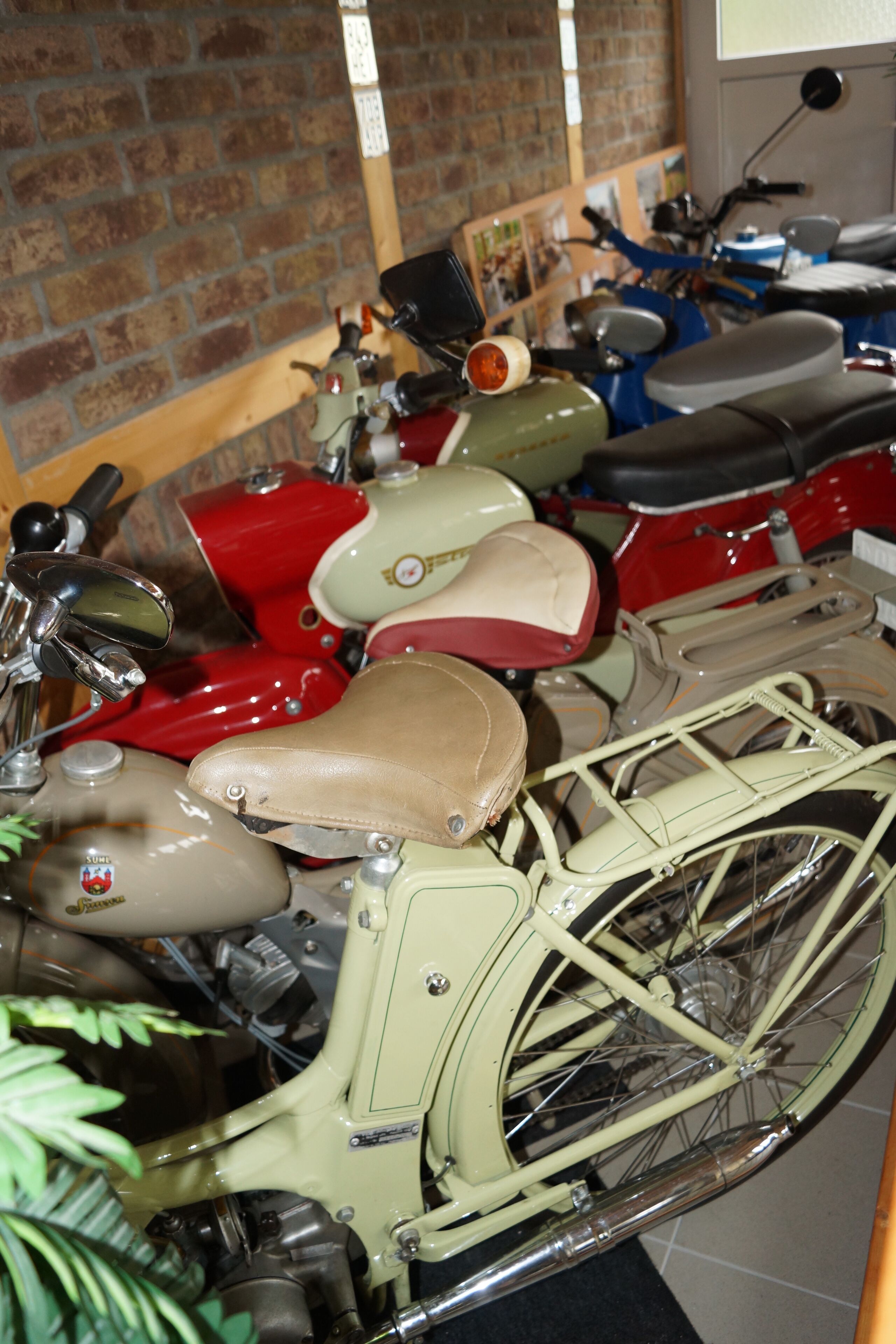 Uma motocicleta antiga com assento bege e carroceria vermelha, exibida em ambiente interno com parede de tijolos ao fundo.