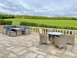 Um pátio com móveis de vime, uma mesa de vidro e vista para um campo verde.