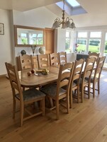 Uma sala de jantar com mesa e cadeiras de madeira, um lustre, um espelho e vista para o verde lá fora.