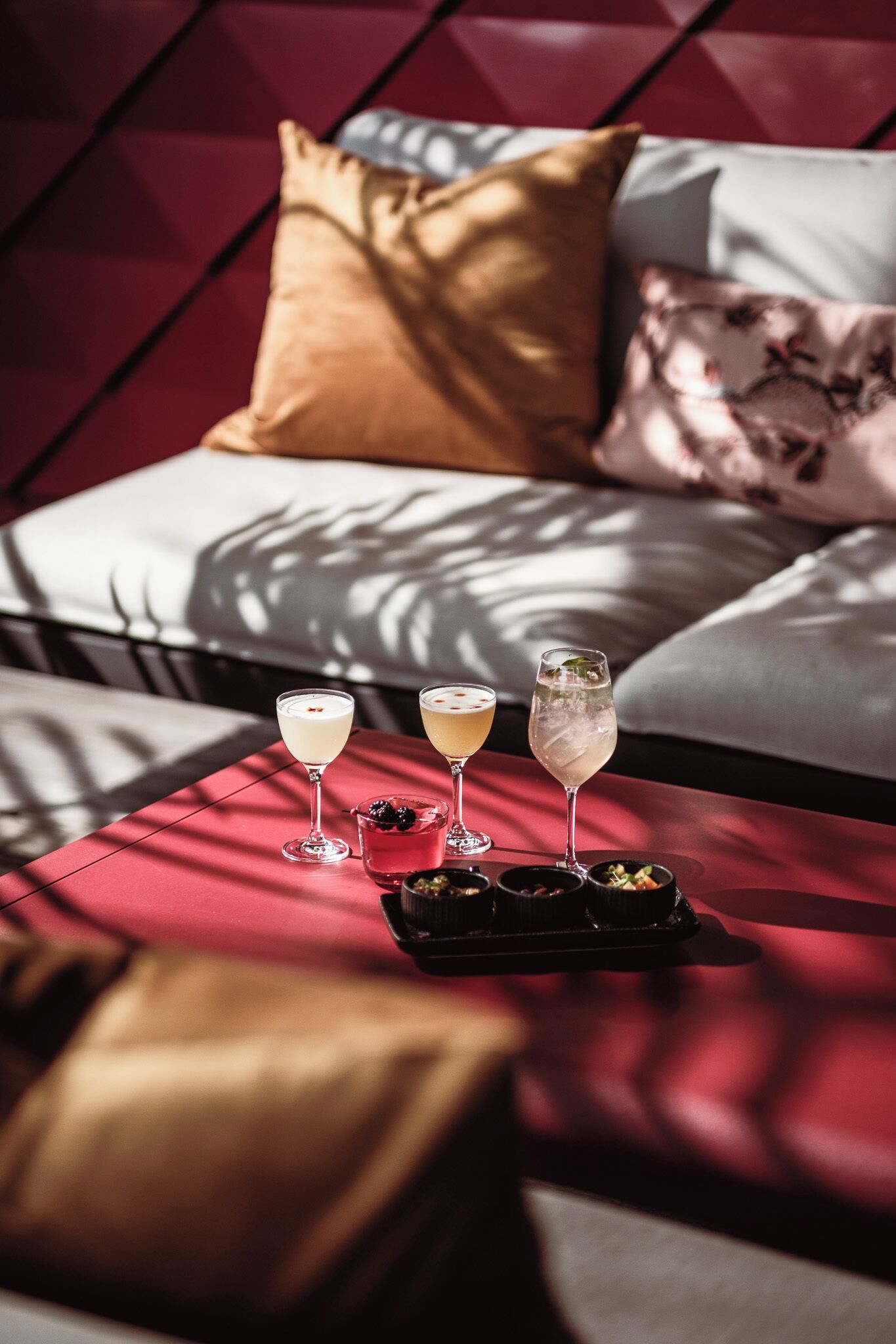 Uma cama com cabeceira vermelha, um travesseiro branco e um travesseiro marrom. Três copos com bebidas estão sobre uma mesa vermelha em frente à cama.