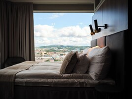 Quarto com cama, almofadas, cabeceira com luzes fixadas na parede, janela com vista para a cidade e cortina.
