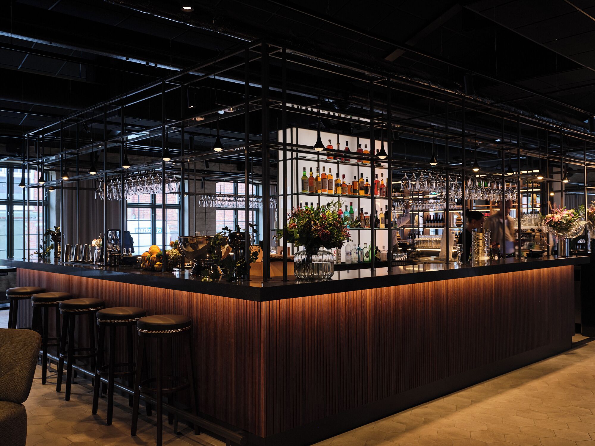 Uma área de bar moderna com balcão de madeira, banquetas e diversos frascos e copos.