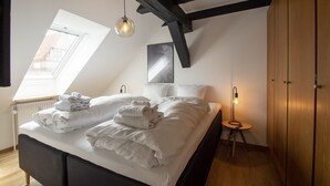 Um quarto com teto inclinado, cama com roupa de cama branca, claraboia, luminária pendente e uma pequena mesa de cabeceira com abajur.