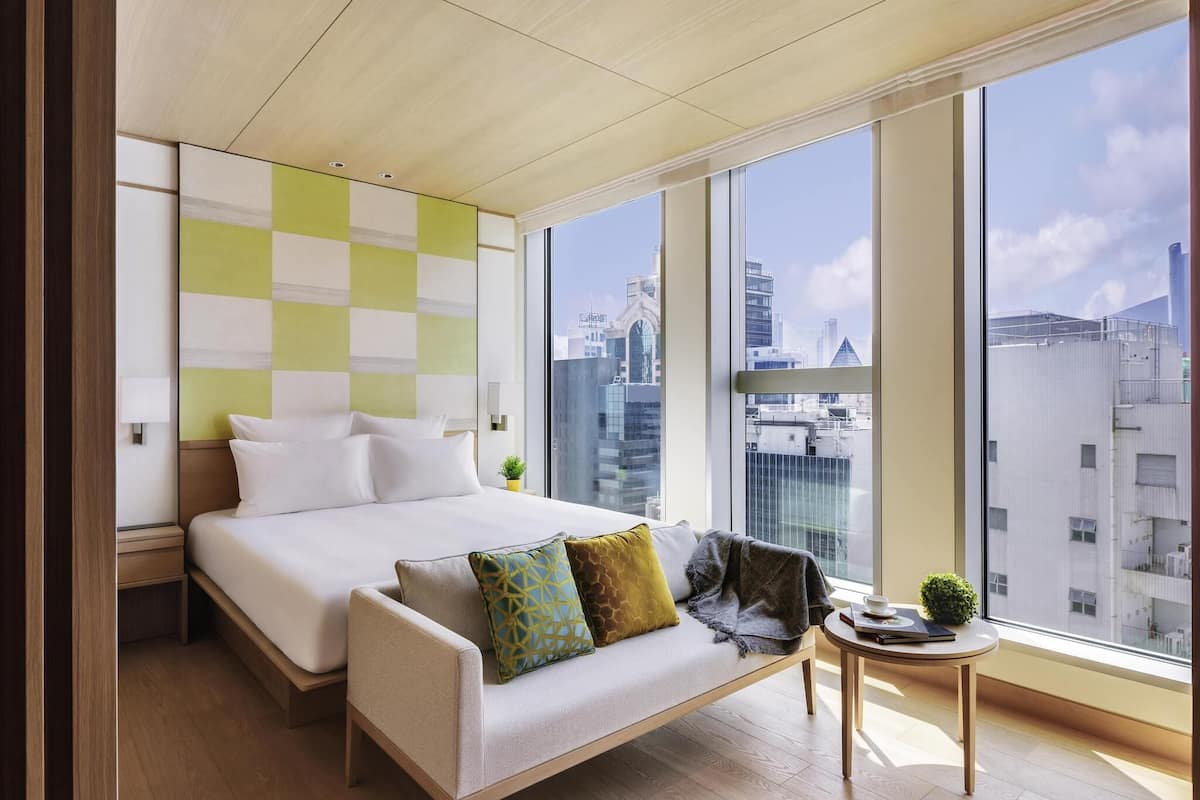 Quarto de hotel moderno com uma cama grande, um sofá, uma mesa pequena e vista para a cidade através de grandes janelas.
