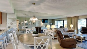 Uma sala de estar moderna com uma mesa de jantar redonda de madeira, cadeiras brancas, um sofá marrom e uma poltrona reclinável. Um espelho grande reflete o ambiente, e há uma TV fixada na parede.