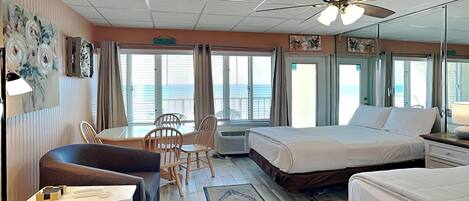 Quarto de hotel com uma cama, uma escrivaninha com cadeira, um sofá, um ventilador de teto e vista para o oceano.