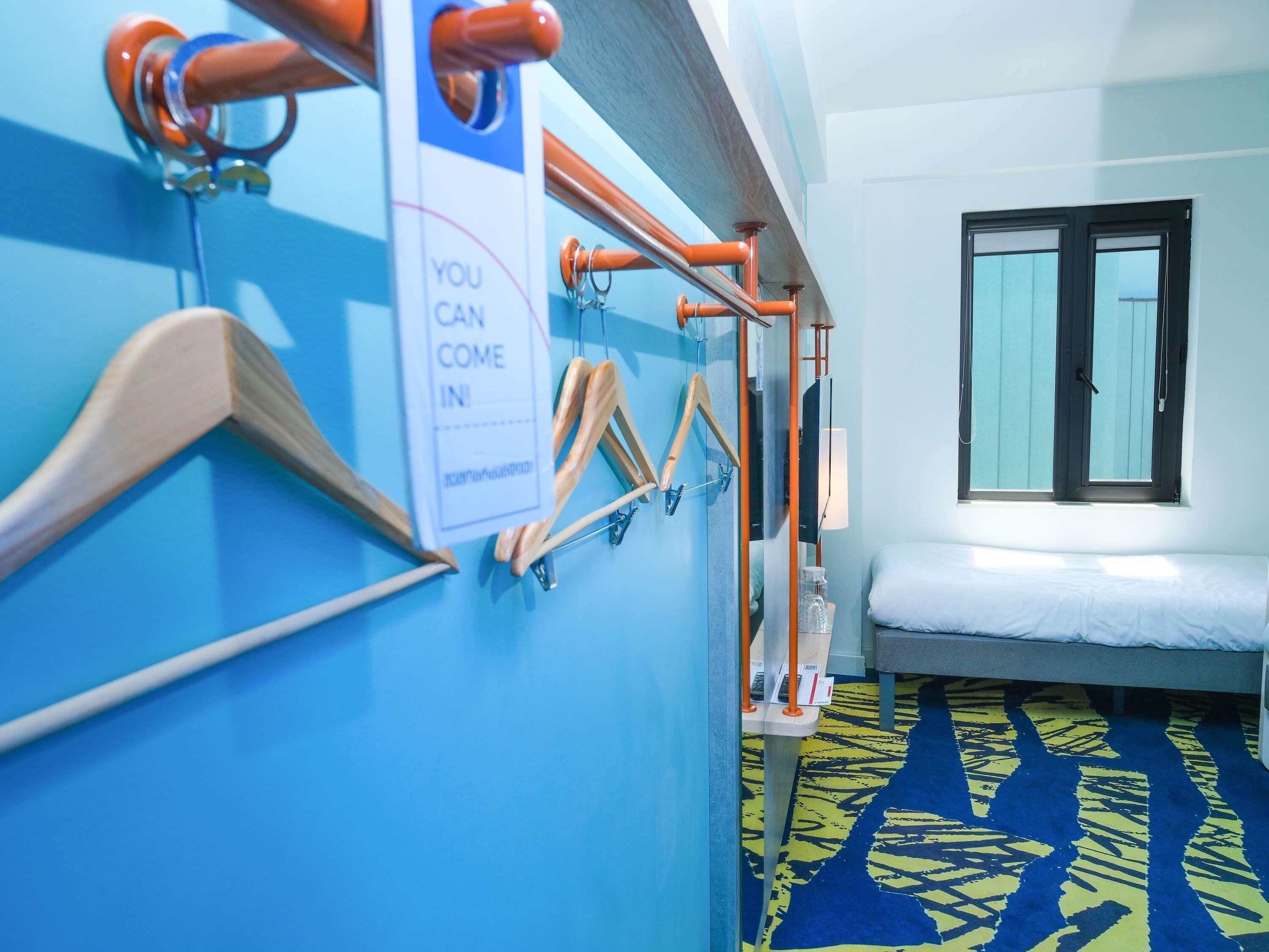 Quarto de hotel com cama, janela, parede azul e um aviso que diz 'PODE ENTRAR!'.