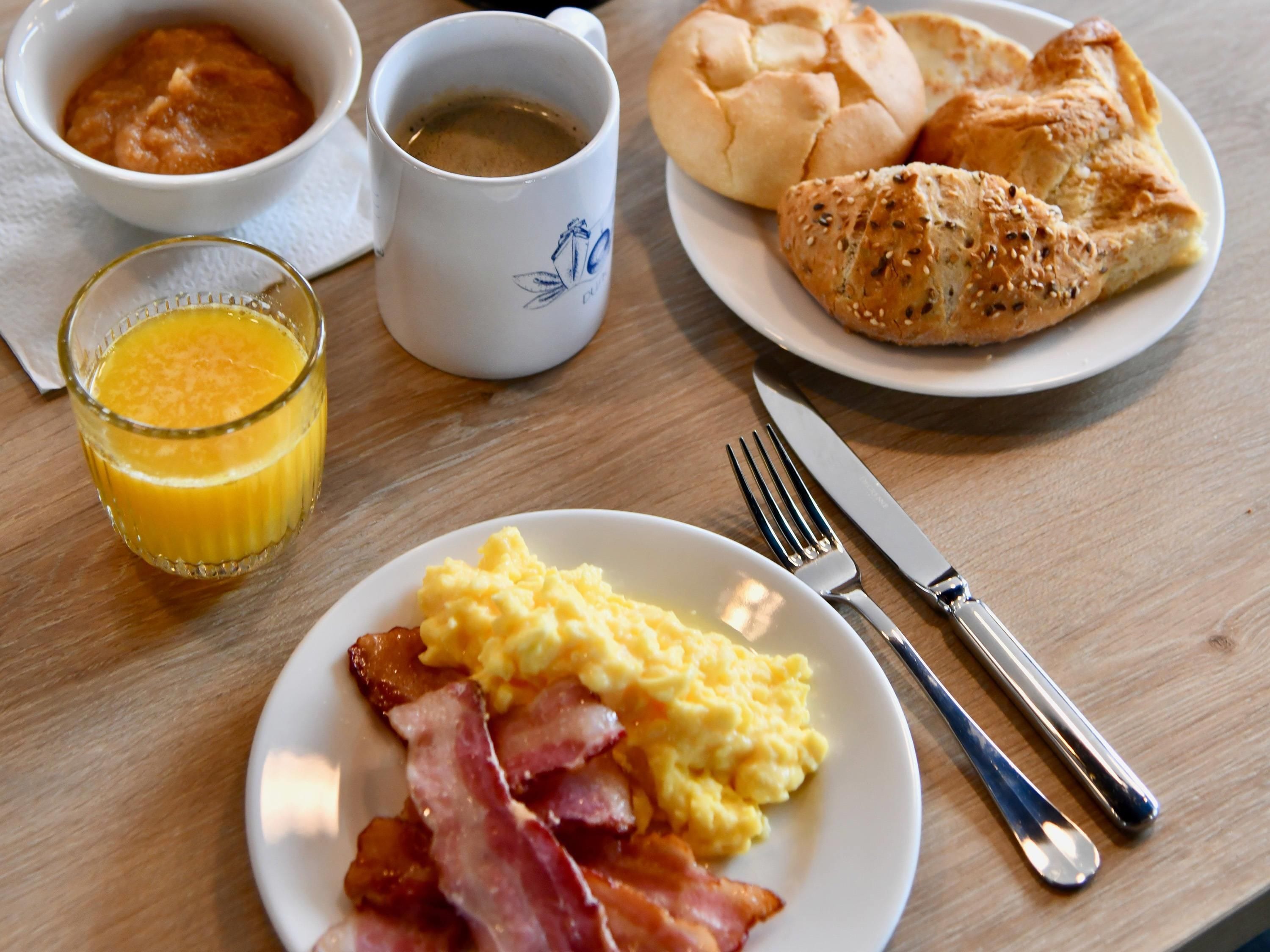 Disposição de pequeno-almoço com ovos mexidos, bacon, tosta, café e sumo de laranja sobre uma mesa de madeira.