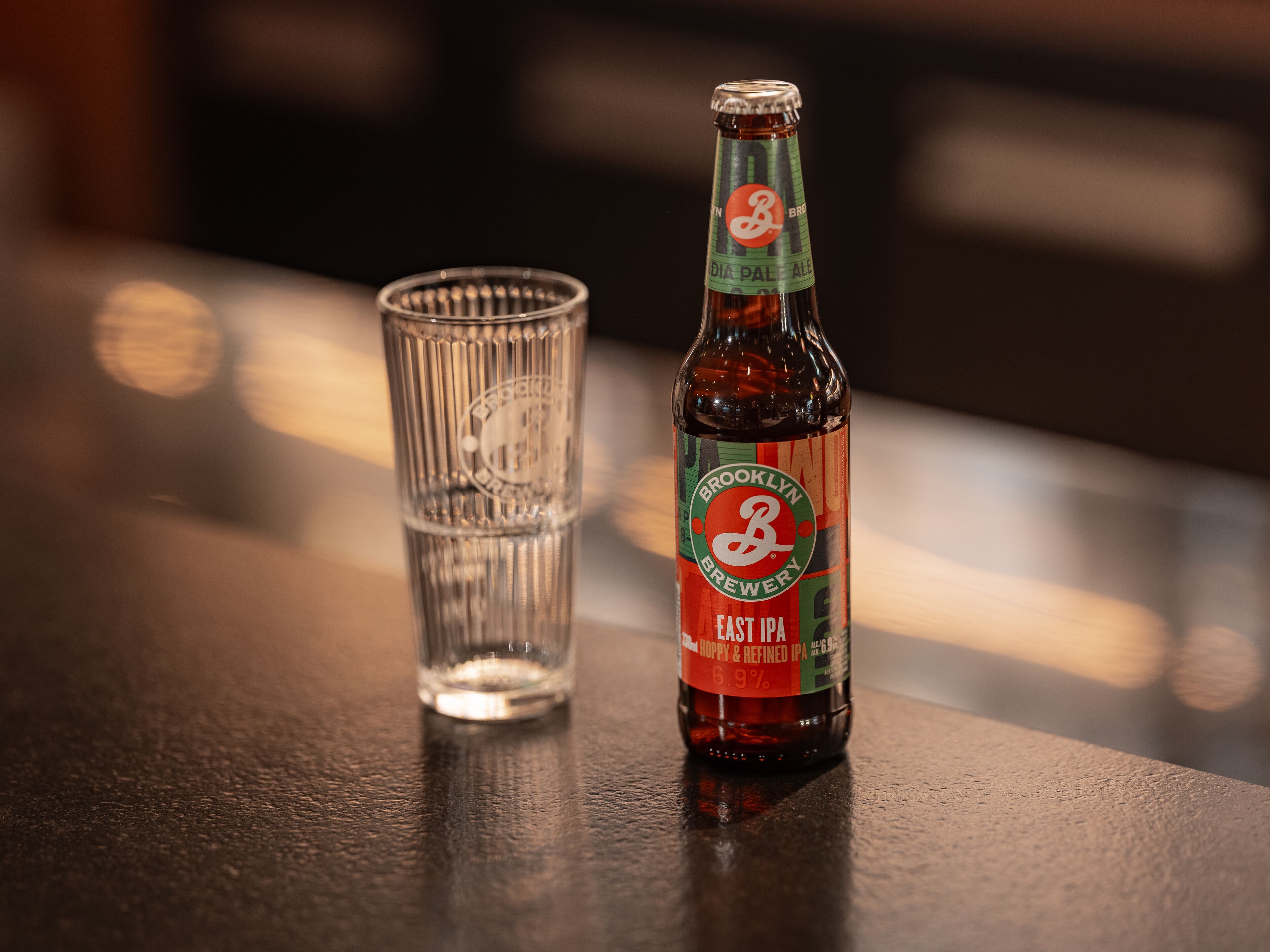 Uma garrafa de Fast IPA da Brooklyn Brewery e um copo de vidro transparente sobre uma superfície escura.