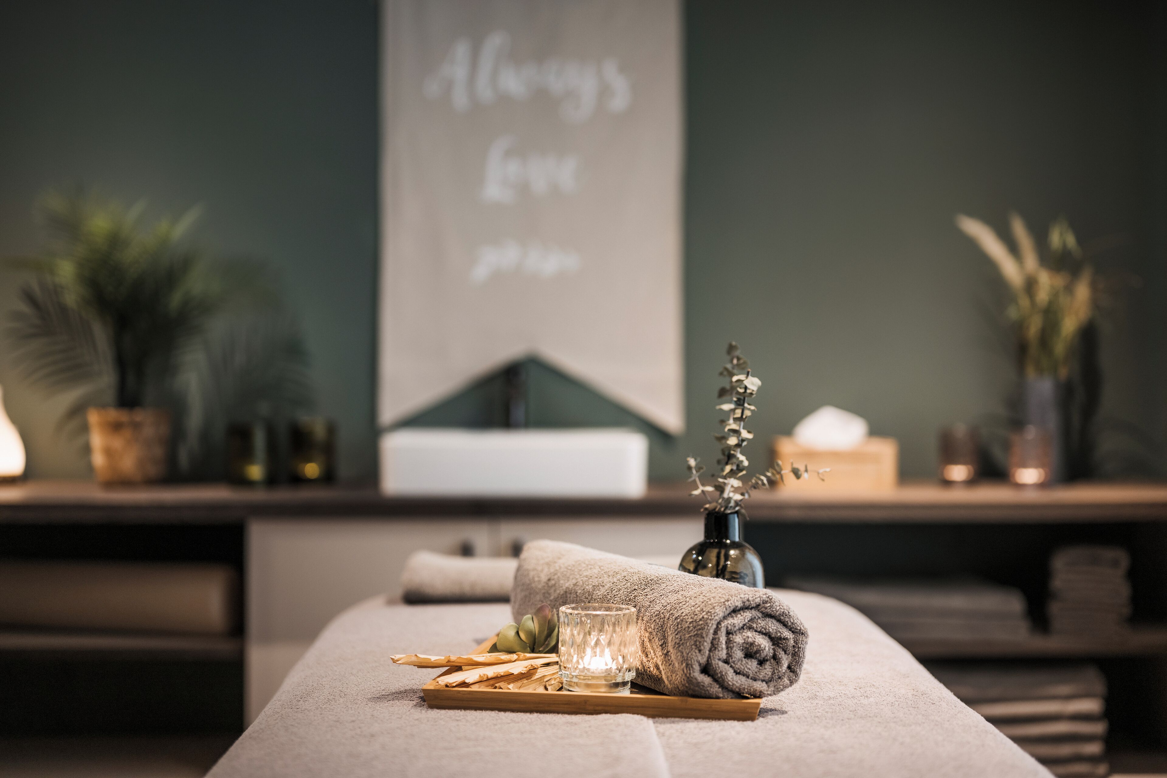 Uma sala de spa com mesa de massagem, toalha enrolada, vela em copo e parede com frase motivacional.