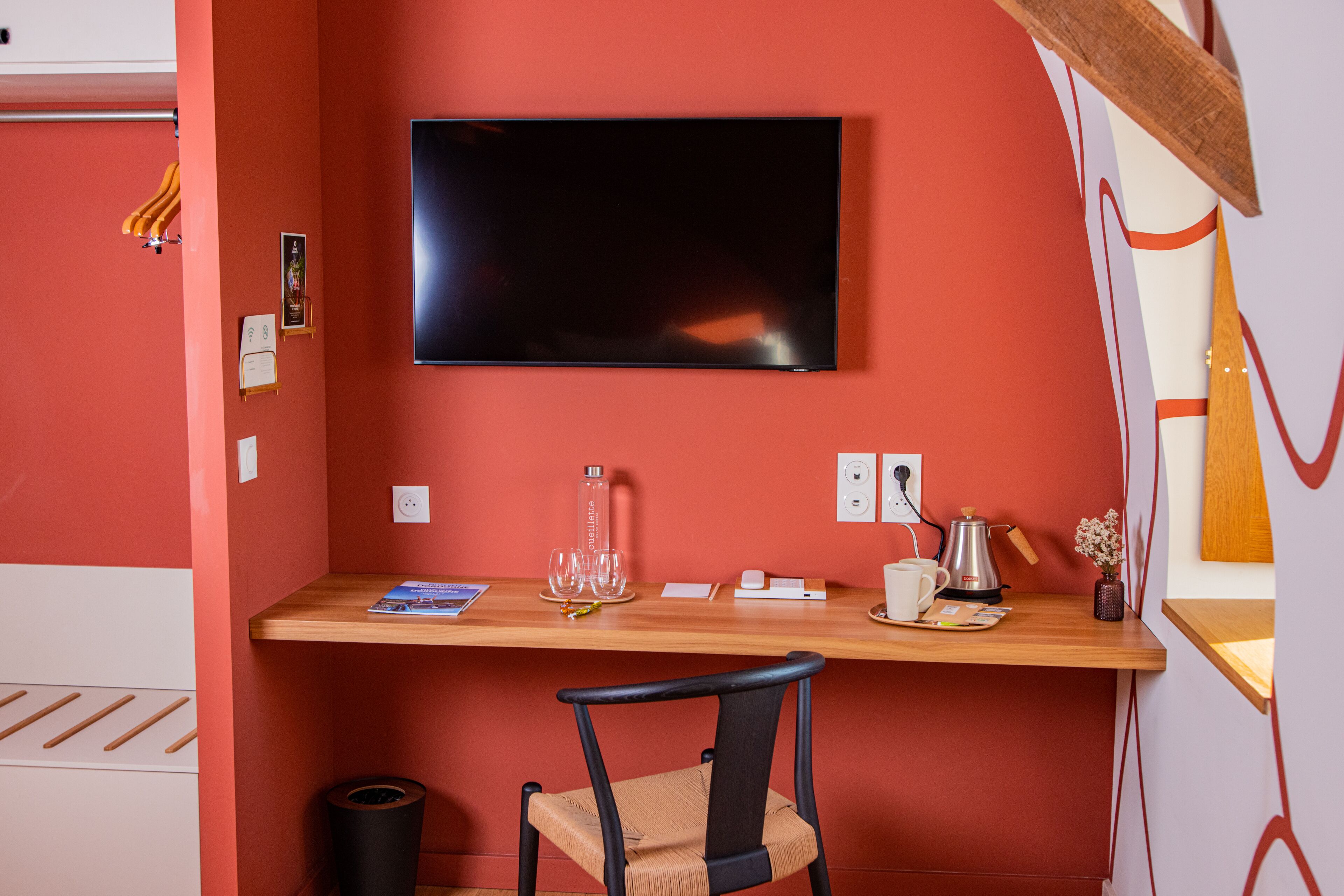 Um quarto com paredes vermelhas, uma escrivaninha de madeira, uma cadeira preta, uma TV fixada na parede e uma pequena lixeira.