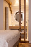 Quarto com cama, vigas de madeira, banheiro com espelho redondo e pia.