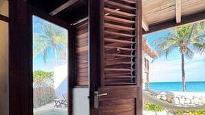 Quarto com porta de madeira, uma pequena mesa redonda com uma toalha, e vista para uma praia com palmeiras e uma piscina.