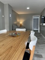 Uma área de refeições com mesa de madeira, cadeiras brancas, uma luminária e uma TV em uma estante.