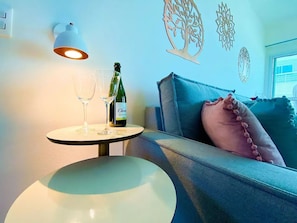 Uma sala de estar aconchegante com uma mesa redonda, uma garrafa de vinho e duas taças. Uma luminária de parede ilumina o ambiente. Arte decorativa na parede e uma janela com vista também são visíveis.