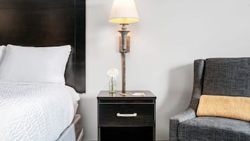 Cama bem arrumada com lençóis brancos, mesa de cabeceira com luminária, pequeno vaso com uma flor e poltrona cinza com almofada amarela.