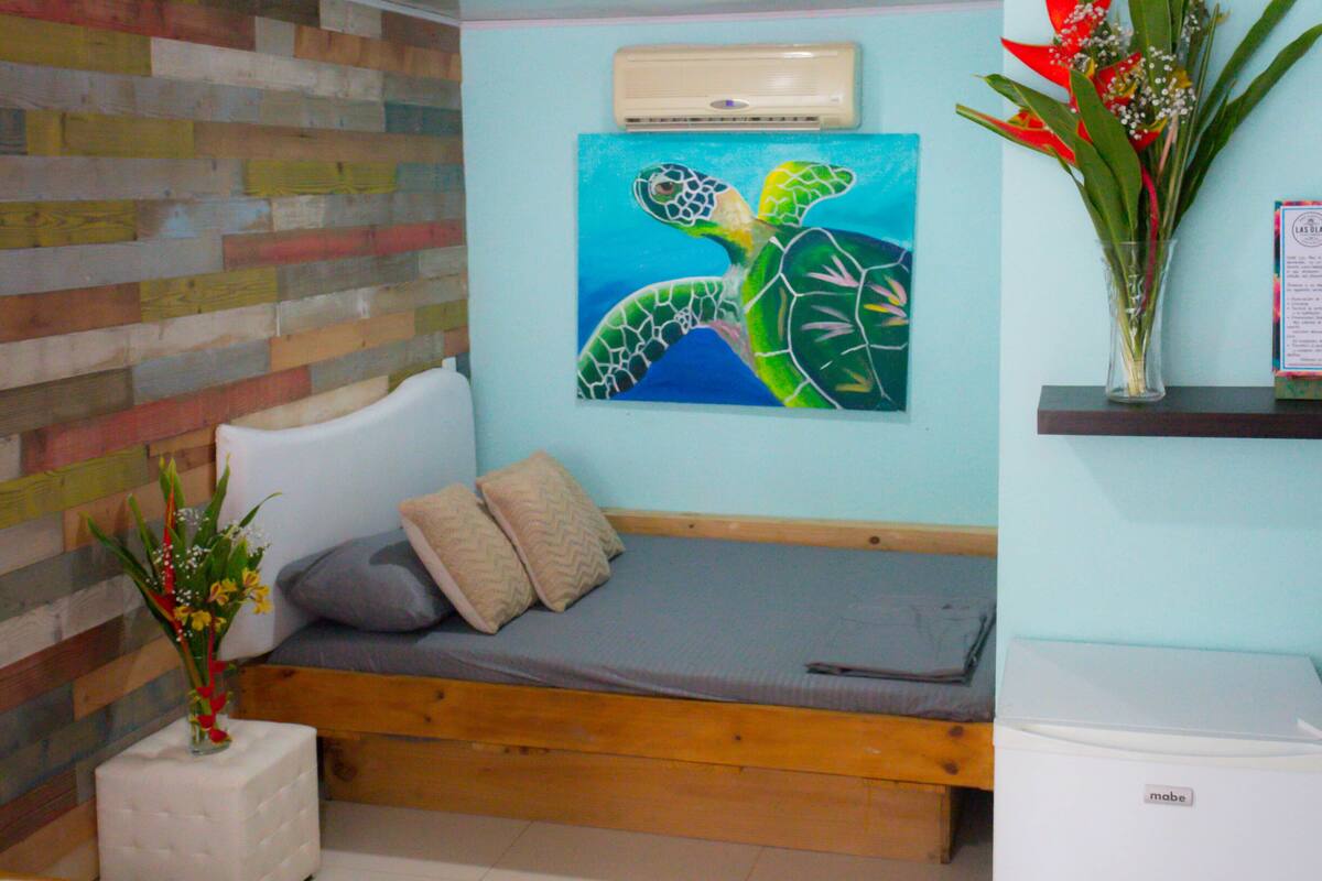Quarto com cama de madeira, ar-condicionado fixado na parede, pintura de uma tartaruga e uma pequena prateleira com um vaso de flores.