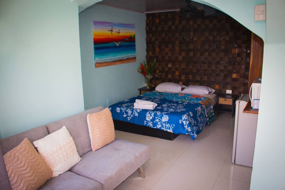 Quarto com cama, sofá, mesa de cabeceira e pequeno espaço de cozinha.
