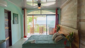 Quarto com cama, ventilador de teto, vista através de porta de vidro para área externa com deck de madeira e cadeiras, e planta em vaso sobre a mesa de cabeceira.