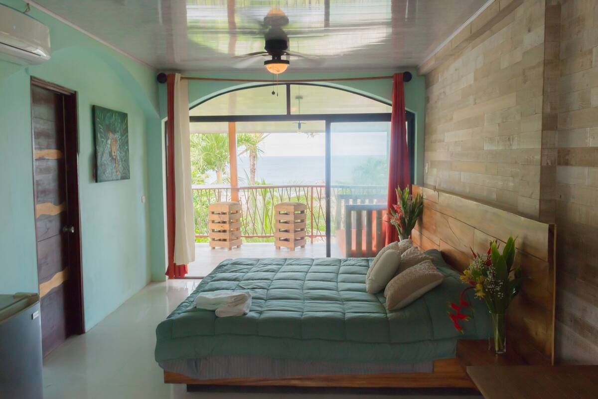 Quarto com cama, ventilador de teto, vista através de porta de vidro para área externa com deck de madeira e cadeiras, e planta em vaso sobre a mesa de cabeceira.