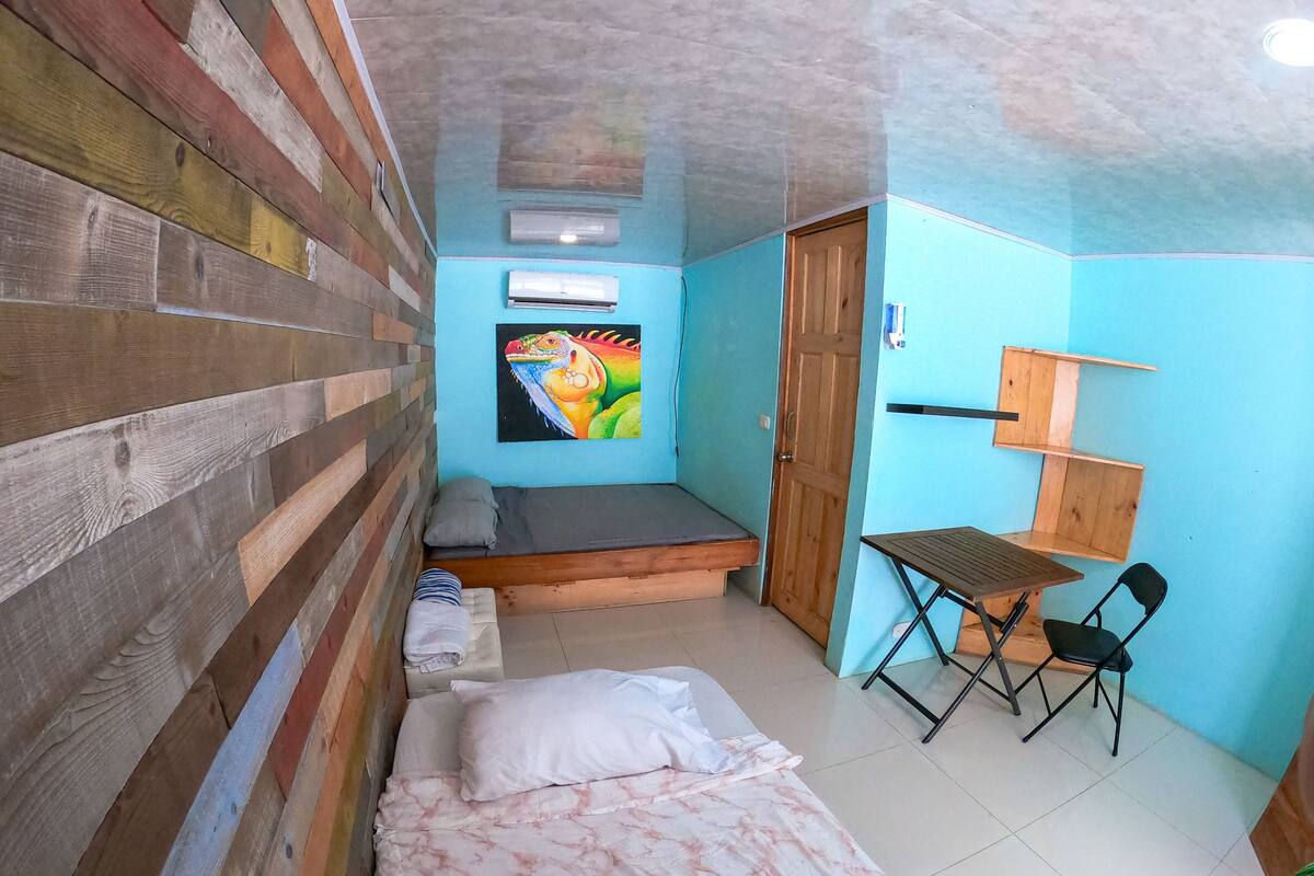 Quarto pequeno e acolhedor com cabeceira de madeira, cama com colcha estampada em rosa e branco, uma escrivaninha com cadeira e um quadro colorido na parede.