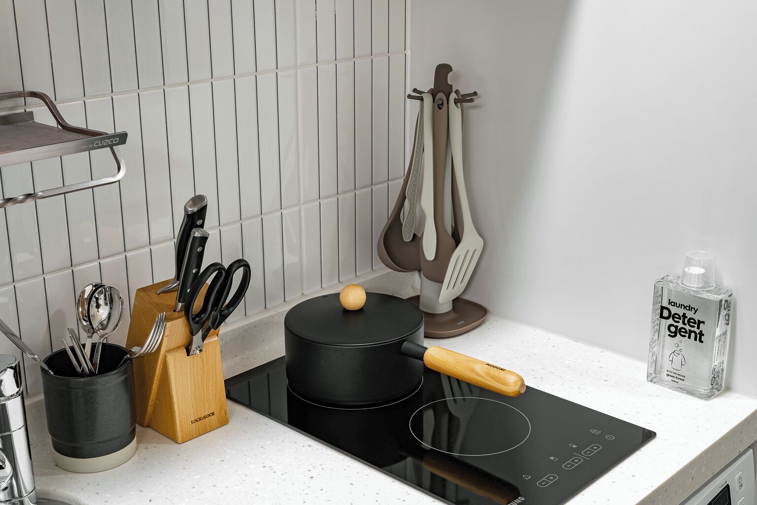 Cozinha com fogão por indução preto, tábua de corte de madeira, panela, porta-facas e uma embalagem de detergente para roupa.